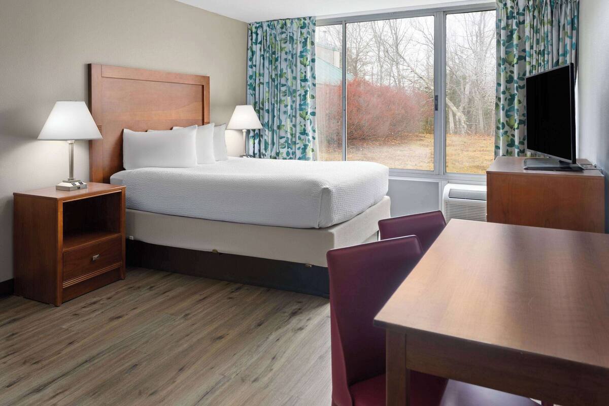 Quarto de hotel com uma cama, duas luminárias de cabeceira, uma TV sobre um suporte de madeira, uma mesa de madeira e uma janela com cortinas.