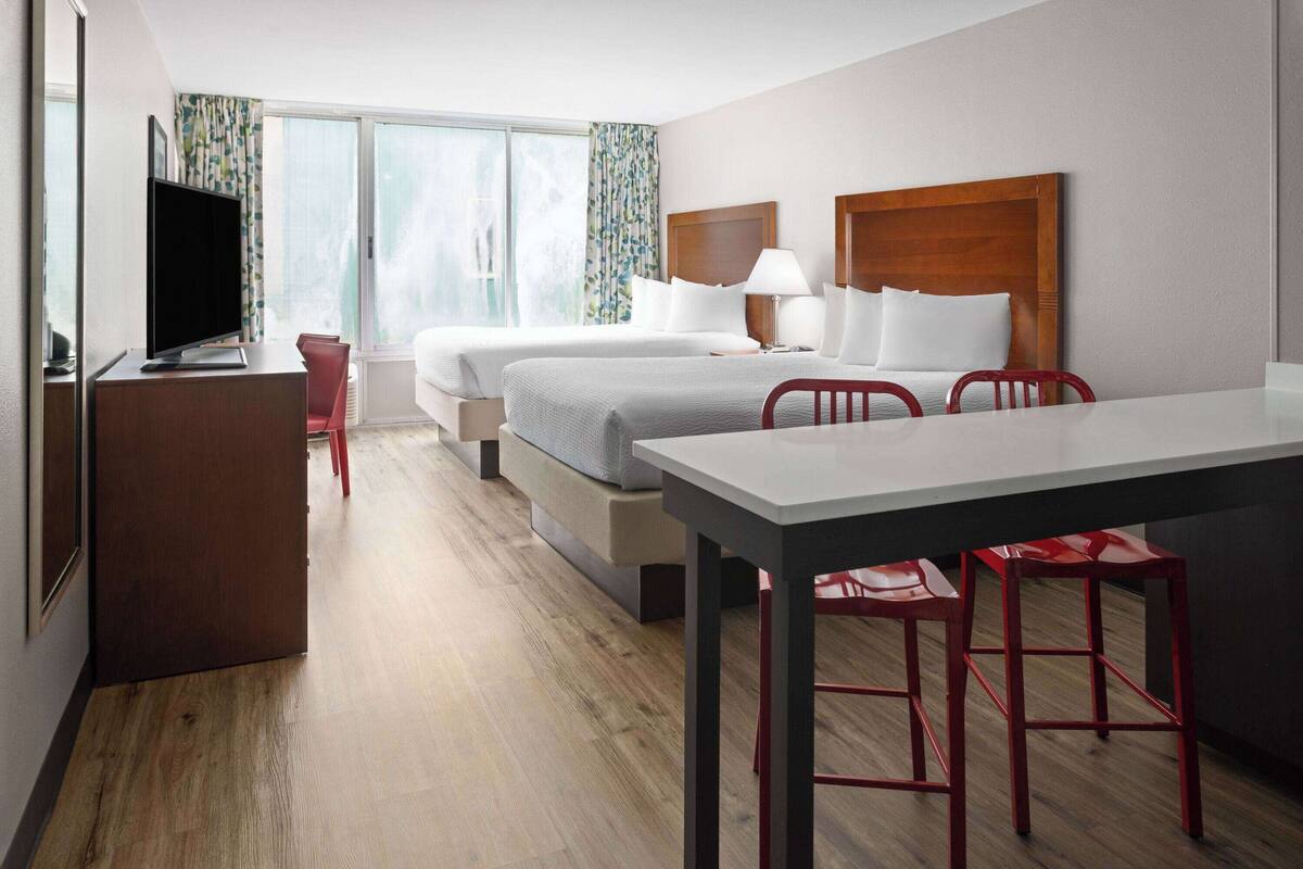 Quarto de hotel com duas camas, uma escrivaninha com cadeira, televisão e janela com cortinas.