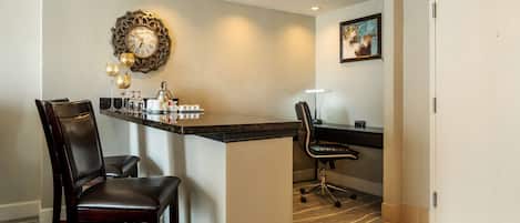 Quarto de hotel com área de jantar, escrivaninha, cadeira, relógio e porta.