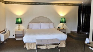 Quarto de hotel bem arrumado, com uma cama grande, duas mesas de cabeceira com abajures, um banco, uma mesinha com um menu e um radiador.