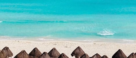 Uma praia com guarda-sóis de palha e espreguiçadeiras, vista clara do oceano e uma costa arenosa.