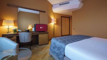 Quarto de hotel com uma cama, uma escrivaninha com computador, uma cadeira e uma luminária.