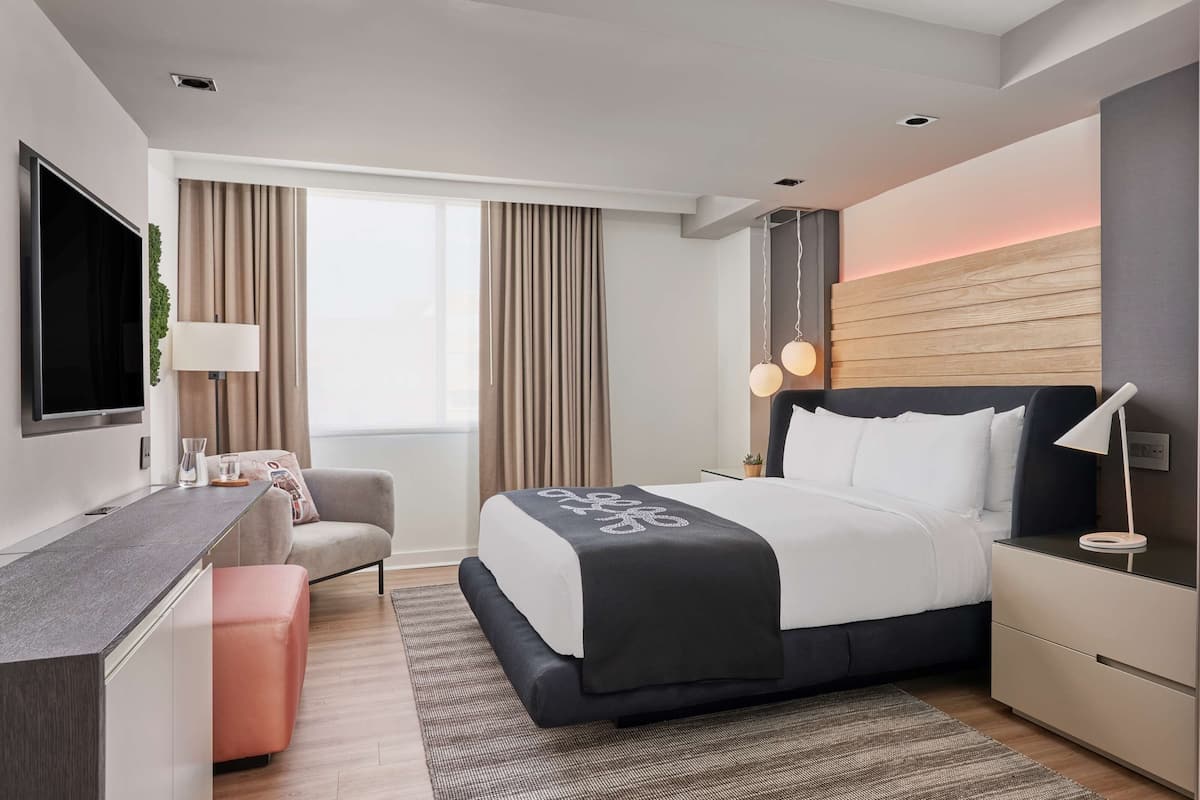 Quarto de hotel moderno com cama, mesa de cabeceira, abajur e escrivaninha com cadeira.