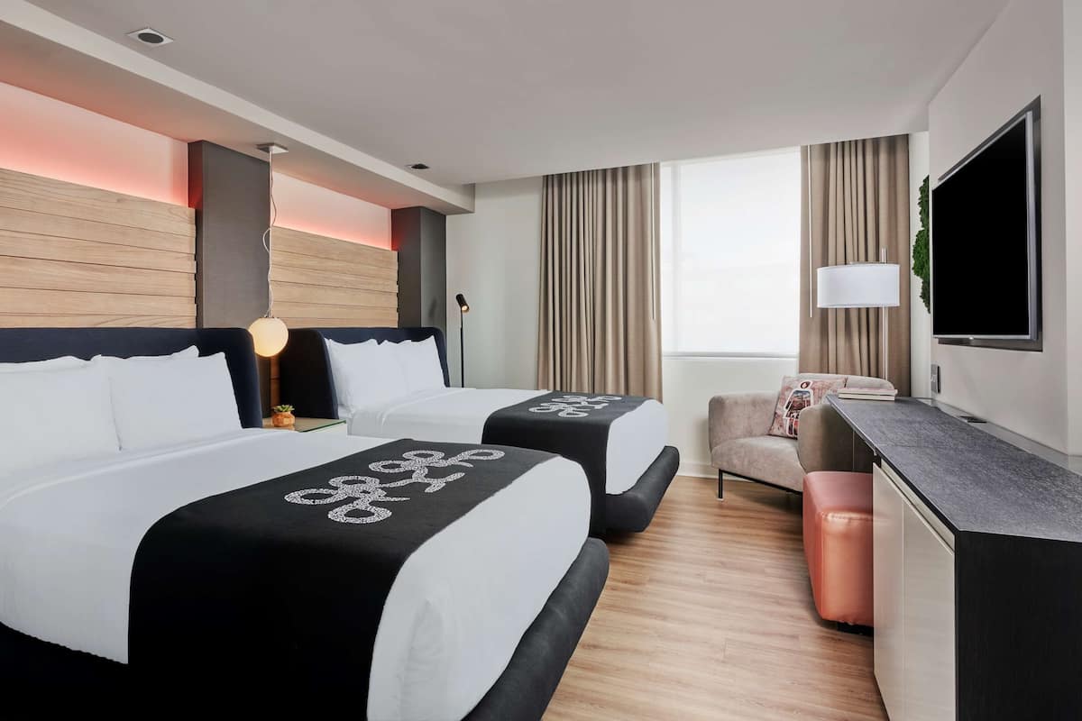 Quarto de hotel com duas camas, uma escrivaninha, televisão e janela com cortinas.