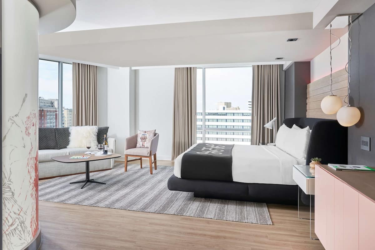 Quarto de hotel moderno com uma cama grande, um sofá, uma mesinha e vista para a cidade.