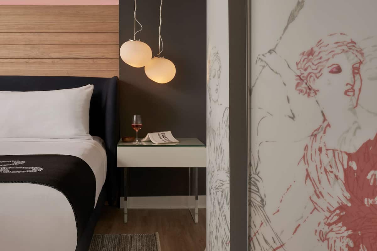 Quarto de hotel moderno com cama, mesa de cabeceira e parede com pintura abstrata vermelha.