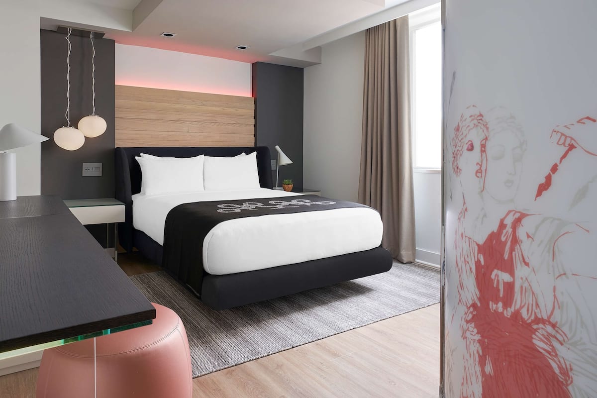 Quarto de hotel moderno com uma cama, mesas de cabeceira, escrivaninha e uma janela grande.