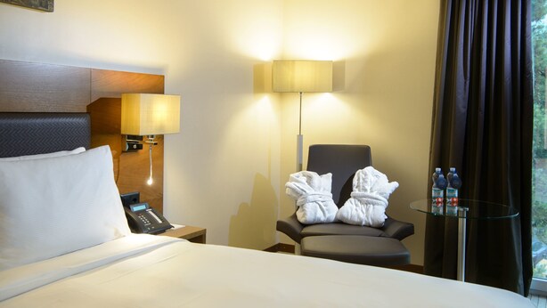 Quarto de hotel com cama, uma cadeira com toalhas, luminária, telefone e janela com cortinas.