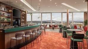 Uma área de bar com vista para a cidade, banquetas e uma mistura de cadeiras vermelhas e verdes.