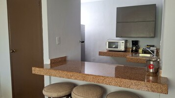 Uma cozinha compacta com micro-ondas, cafeteira e uma pia pequena. Há três bancos sob o balcão.