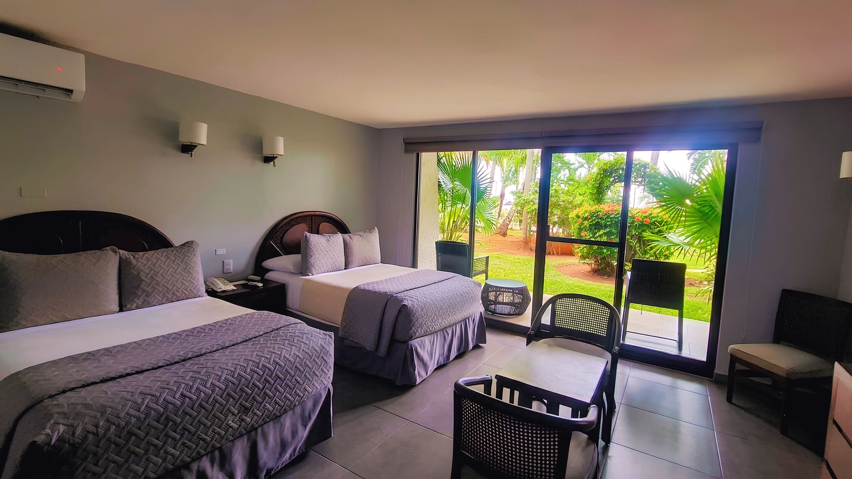 Quarto de hotel com duas camas, uma escrivaninha, cadeiras e uma grande porta de vidro deslizante para um jardim.