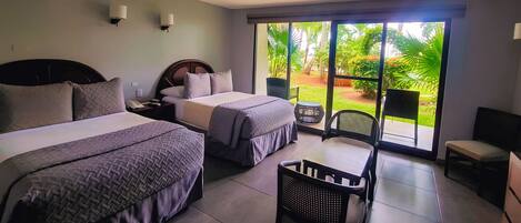Quarto de hotel com duas camas, uma escrivaninha, cadeiras e uma grande porta de vidro deslizante para um jardim.