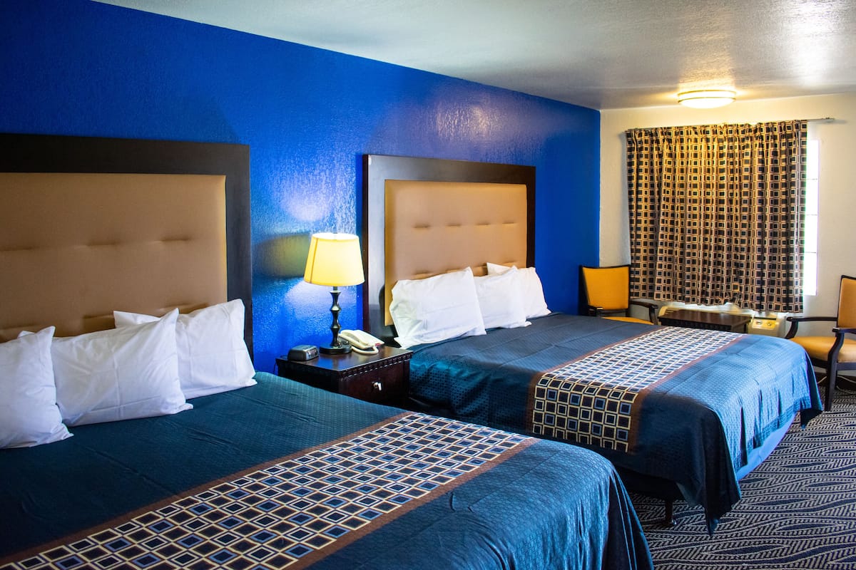 Quarto de hotel com duas camas, uma escrivaninha, uma cadeira, um abajur e uma janela com cortinas.