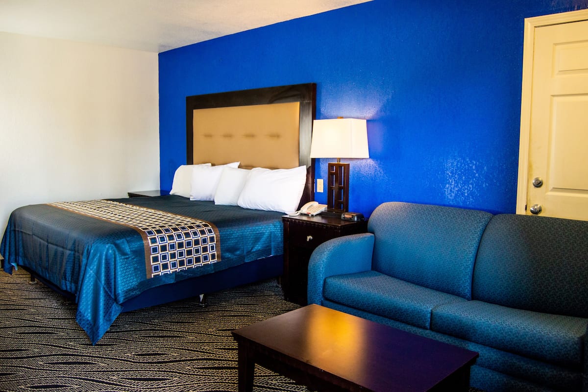 Quarto de hotel com cama, sofá, mesa de cabeceira com abajur e piso coberto por carpete.
