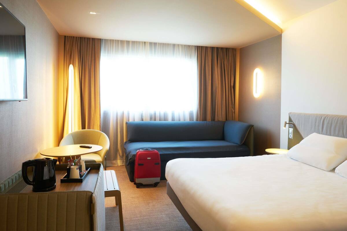 Quarto de hotel com cama, sofá, escrivaninha com cafeteira, mala e janela com cortinas.