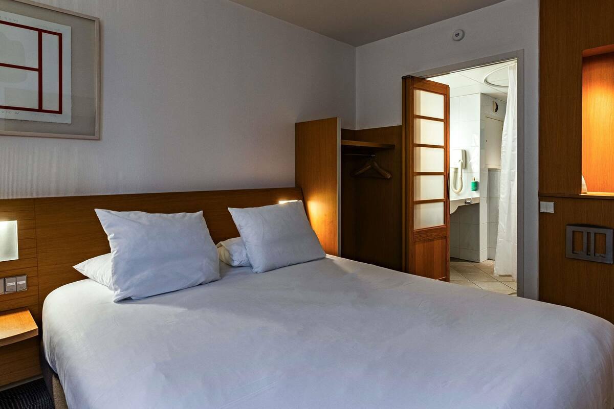 Quarto de hotel com uma cama grande, cabeceira de madeira, mesa de cabeceira com abajur e guarda-roupa com espelho.