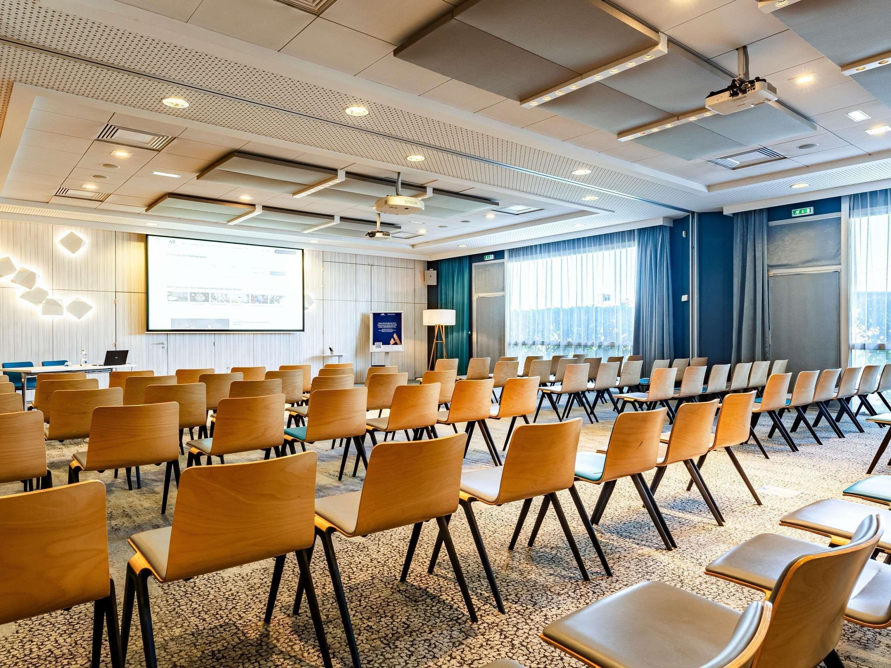 Uma sala de conferência com fileiras de cadeiras, uma tela de projetor e uma área para apresentações.