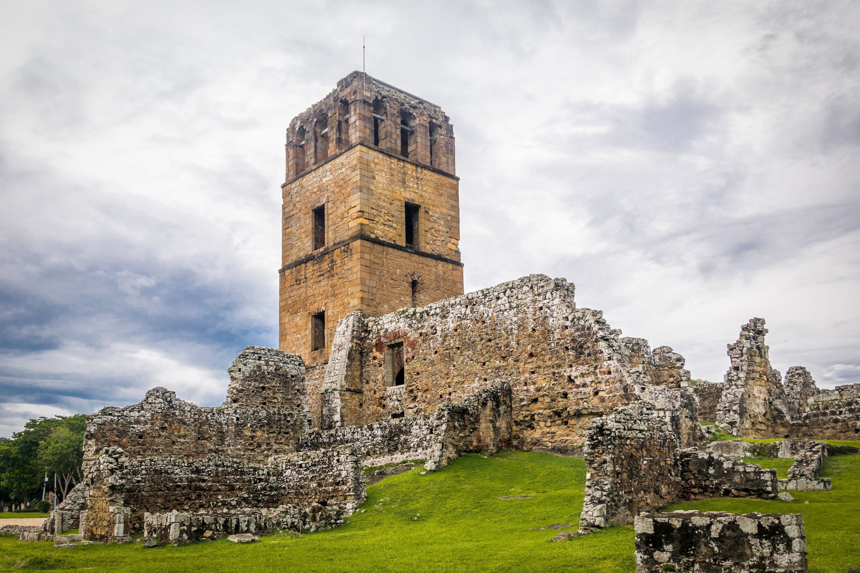Uma torre de pedra histórica com base de tijolos, rodeada por ruínas e áreas ajardinadas.