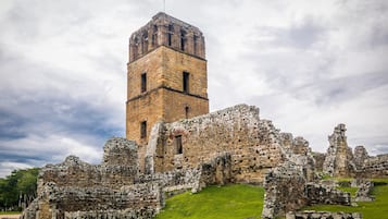 Uma torre de pedra histórica com base de tijolos, rodeada por ruínas e áreas ajardinadas.