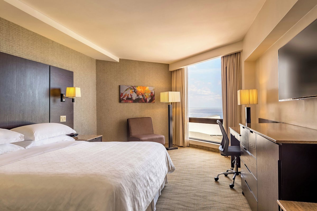 Quarto de hotel com uma cama grande, uma escrivaninha, uma cadeira e uma televisão.