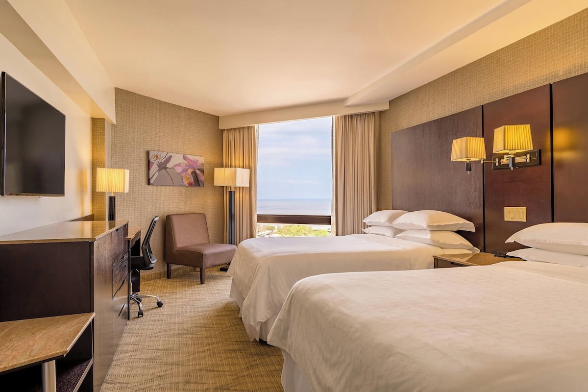 Quarto de hotel com duas camas, uma escrivaninha, uma cadeira, uma TV e uma janela grande com cortinas.