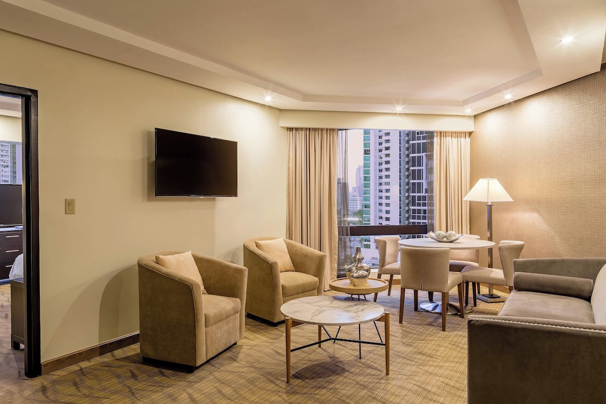 Quarto de hotel com televisão de tela plana, poltronas bege, mesa de centro redonda e vista para prédios altos através de janelas amplas.