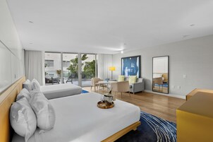 Quarto de hotel com duas camas, uma escrivaninha, uma cadeira, um abajur e uma janela grande com vista para o exterior.
