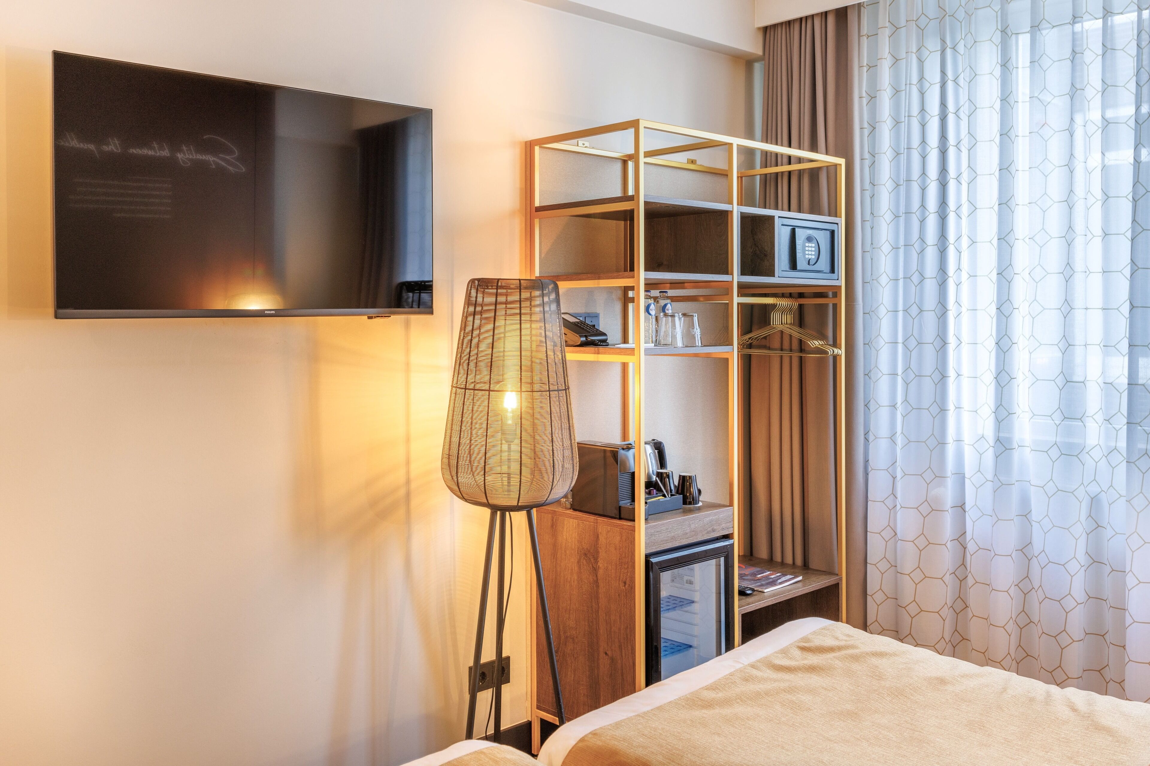 Quarto de hotel com cama, televisão na parede, escrivaninha com luminária, guarda-roupa e janela com cortinas estampadas.