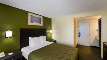 Quarto de hotel com cama, mesa de cabeceira, luminária na parede, um quadro e porta para o banheiro.