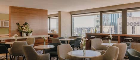 Um hall de hotel com mesas e cadeiras redondas, um balcão de recepção e grandes janelas com vista para a cidade.