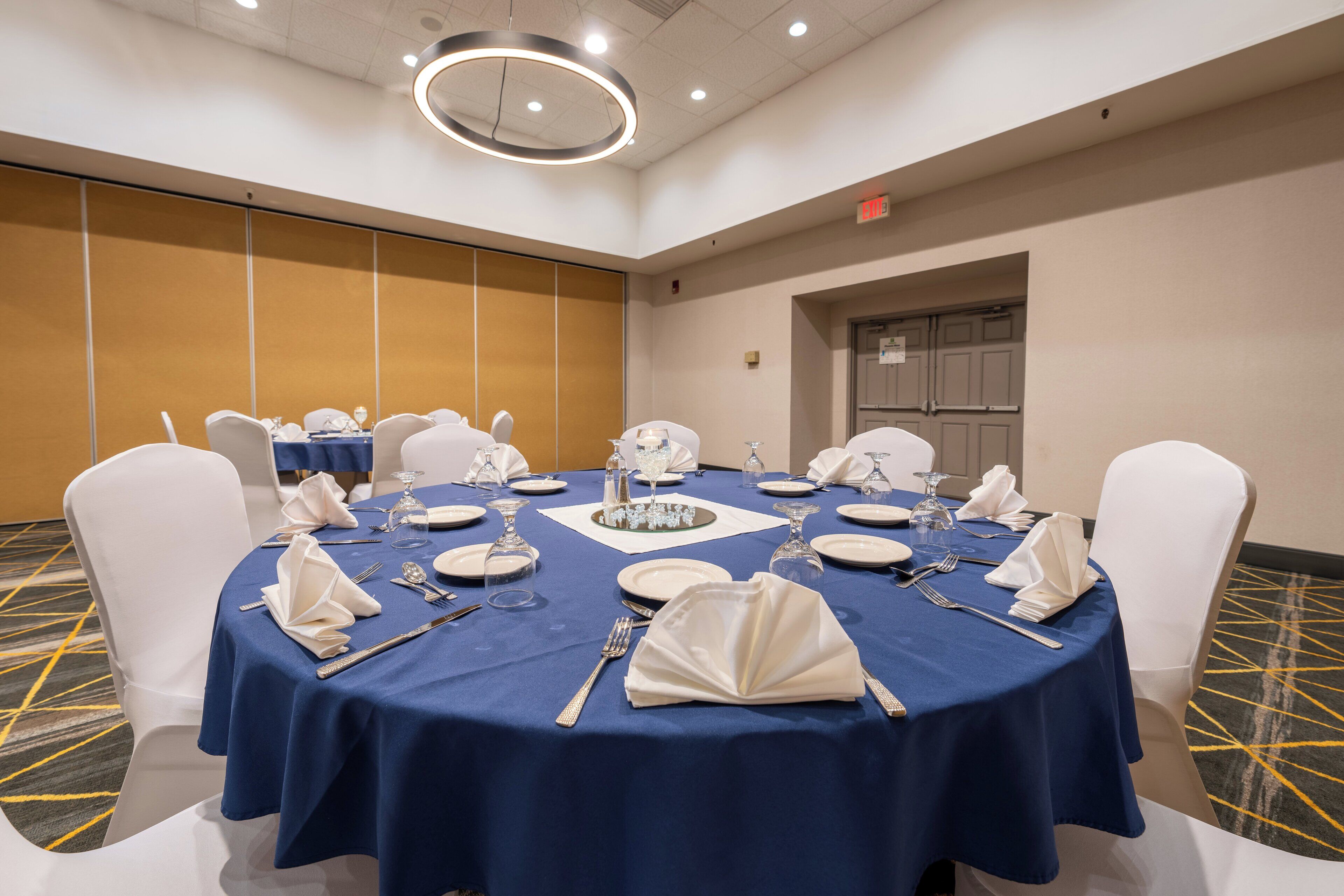 Sala de conferências com mesas redondas preparadas para uma reunião, equipada com placas de identificação, copos d'água e talheres.