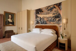 Quarto de hotel com uma cama grande, cabeceira com pintura, mesa de cabeceira com abajur e um banquinho.