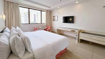 Quarto de hotel com uma cama grande, uma escrivaninha, uma cadeira, uma televisão e uma janela com cortinas.