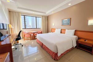 Quarto de hotel com uma cama grande, uma escrivaninha, uma cadeira, uma mesinha e uma janela com cortinas.