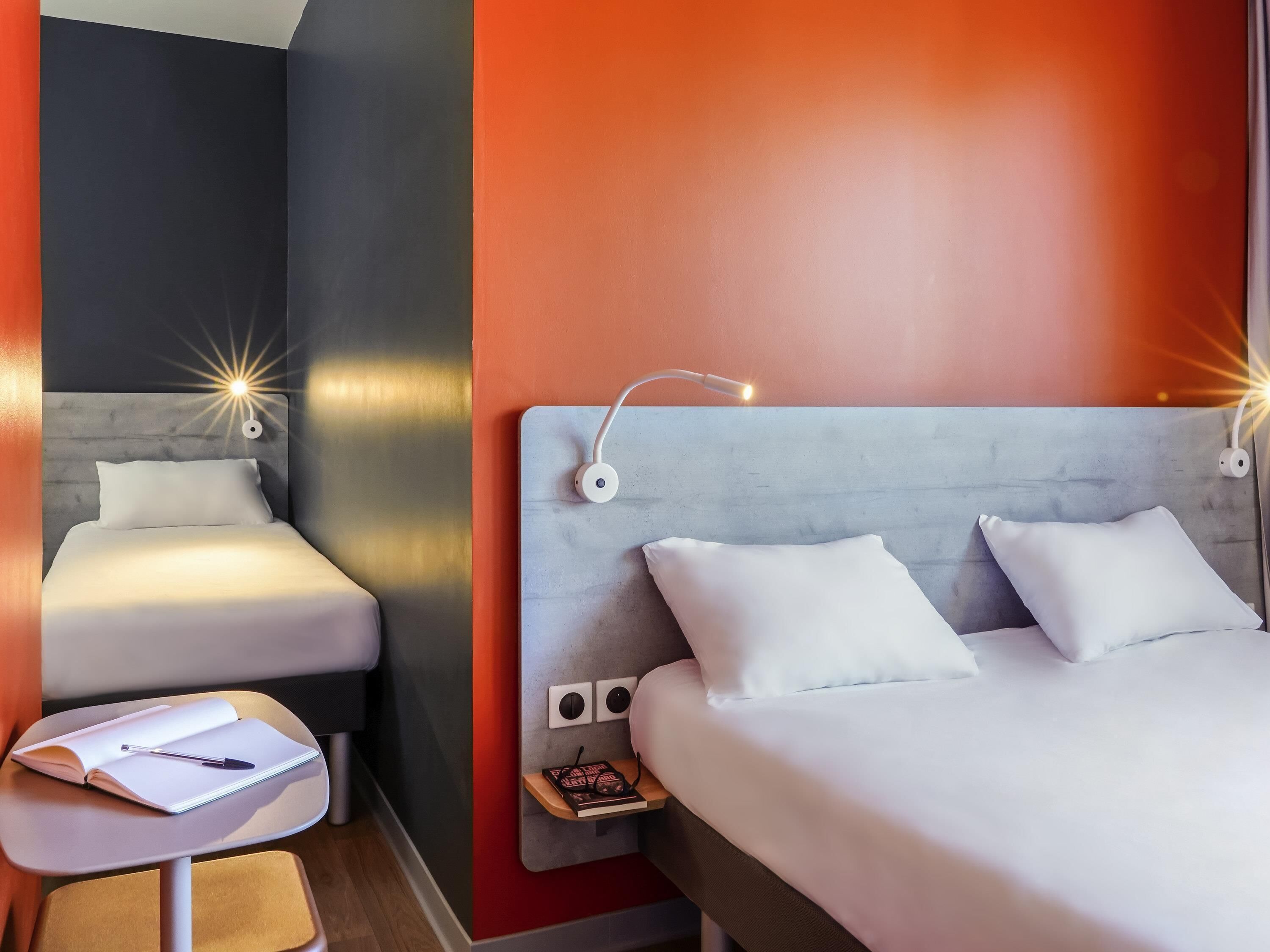 Quarto de hotel com duas camas, uma mesinha com um caderno e caneta, e uma mesa de cabeceira com um livro.