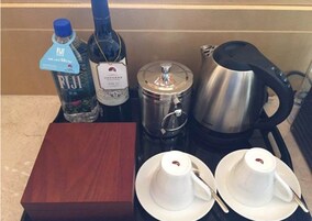 Uma mesa com uma garrafa de água Fiji, uma garrafa de vinho, uma cafeteira, duas xícaras brancas com pires e uma caixa de madeira.