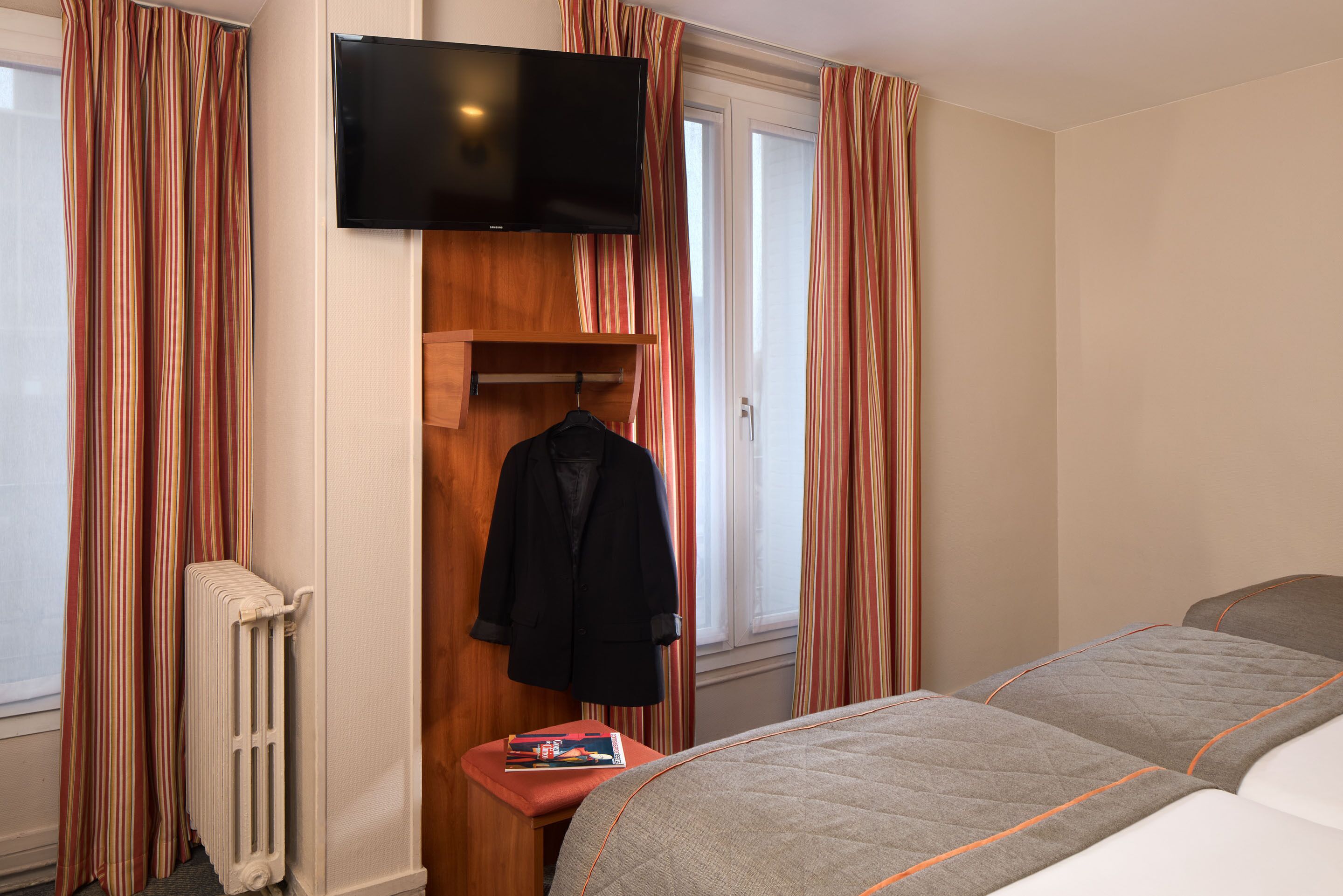 Quarto de hotel com cama, televisão na parede, cabideiro com casaco e cortinas listradas.