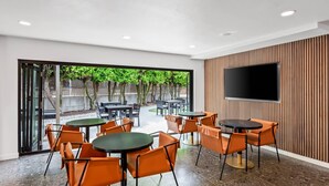 Uma área de estar interna moderna com várias mesas e cadeiras, uma grande TV de tela plana na parede e vista para o exterior por meio de grandes portas de vidro.