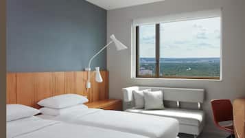 Quarto de hotel com duas camas, uma escrivaninha, uma cadeira, mesa de cabeceira, abajur e uma janela grande com vista para a cidade.