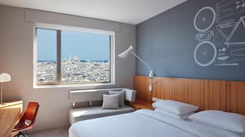 Quarto de hotel com uma grande janela que oferece vista para a cidade, uma cama com roupa de cama branca, uma cabeceira de madeira, uma mesa com cadeira e uma parede com um mural de tema de bicicleta.