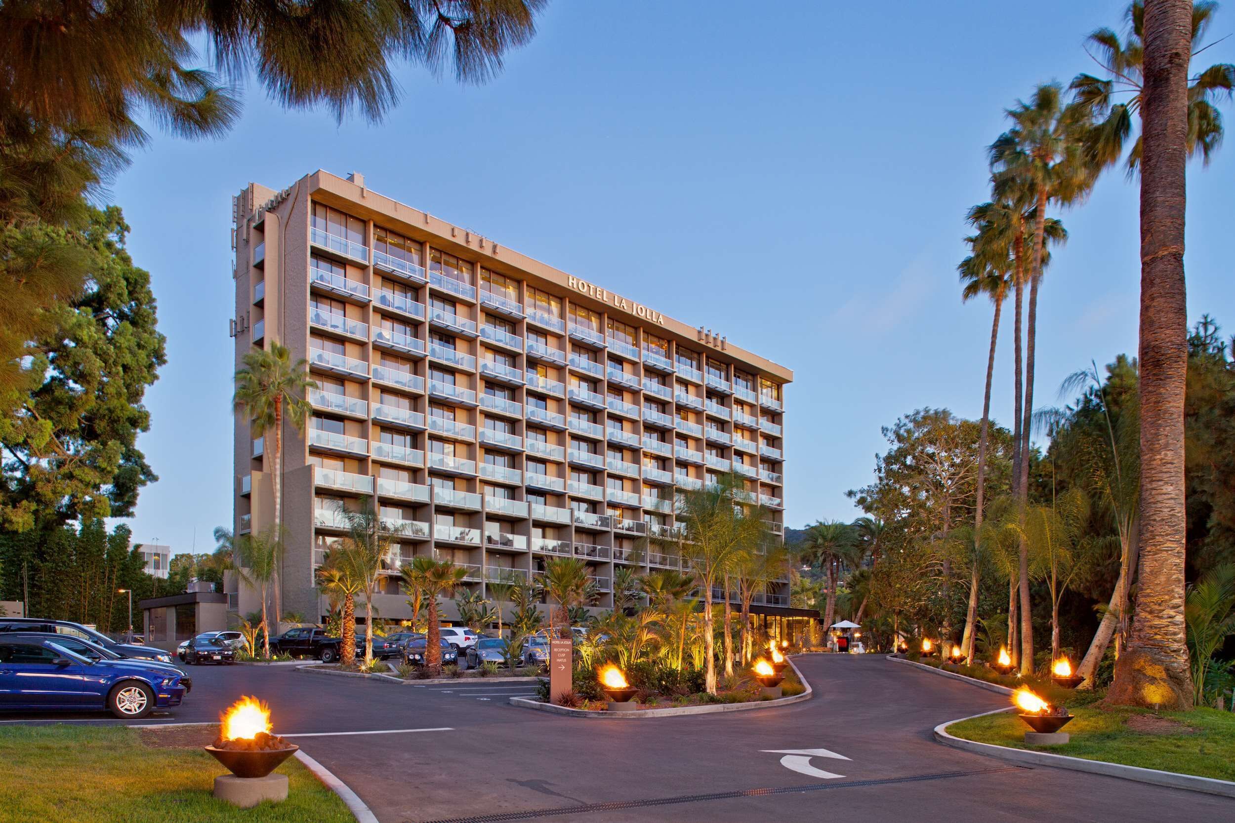 Um hotel com vários andares, um pátio central, palmeiras e céu claro.