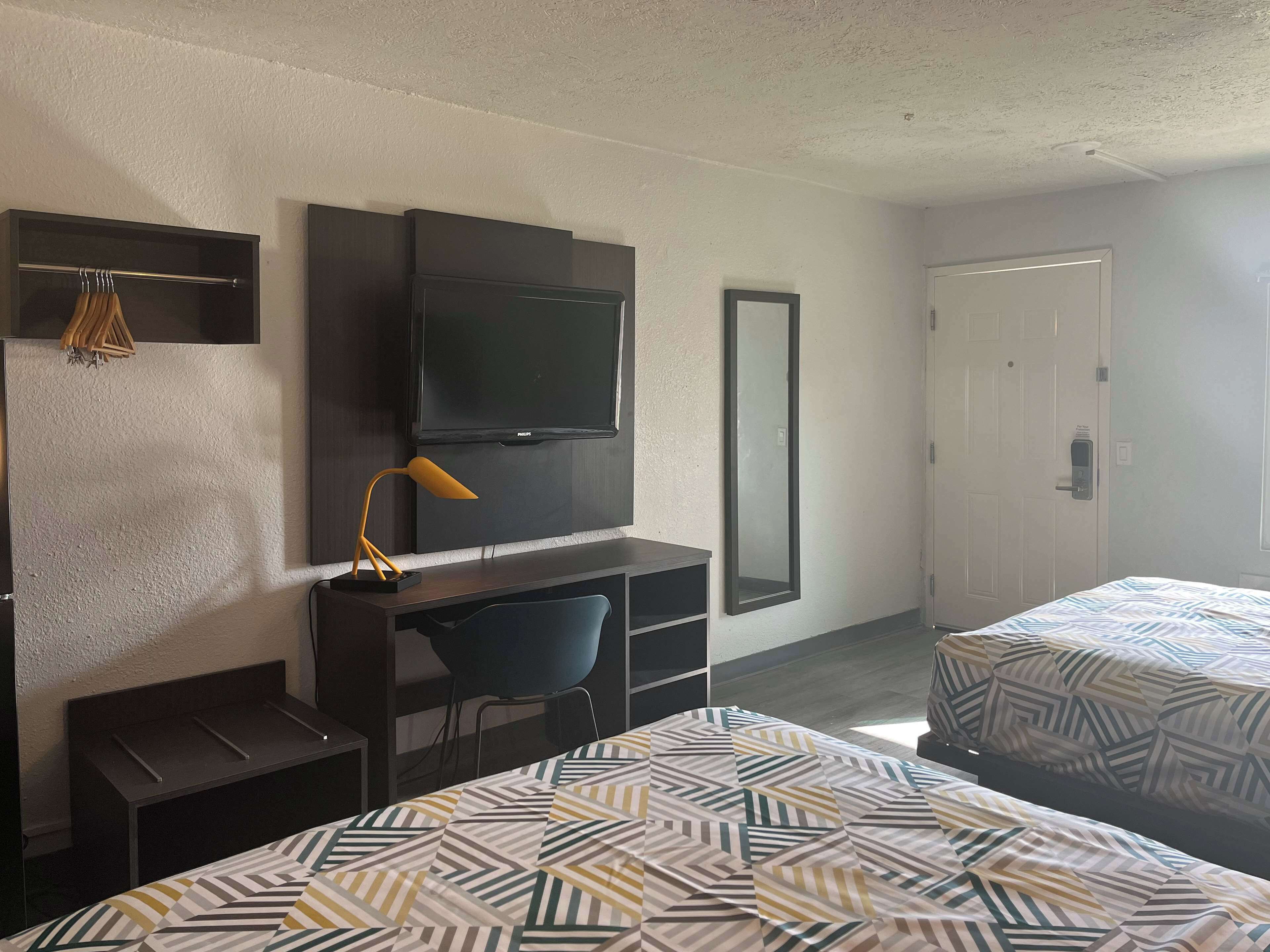 Quarto de hotel com cama, escrivaninha com cadeira, televisão e guarda-roupa.