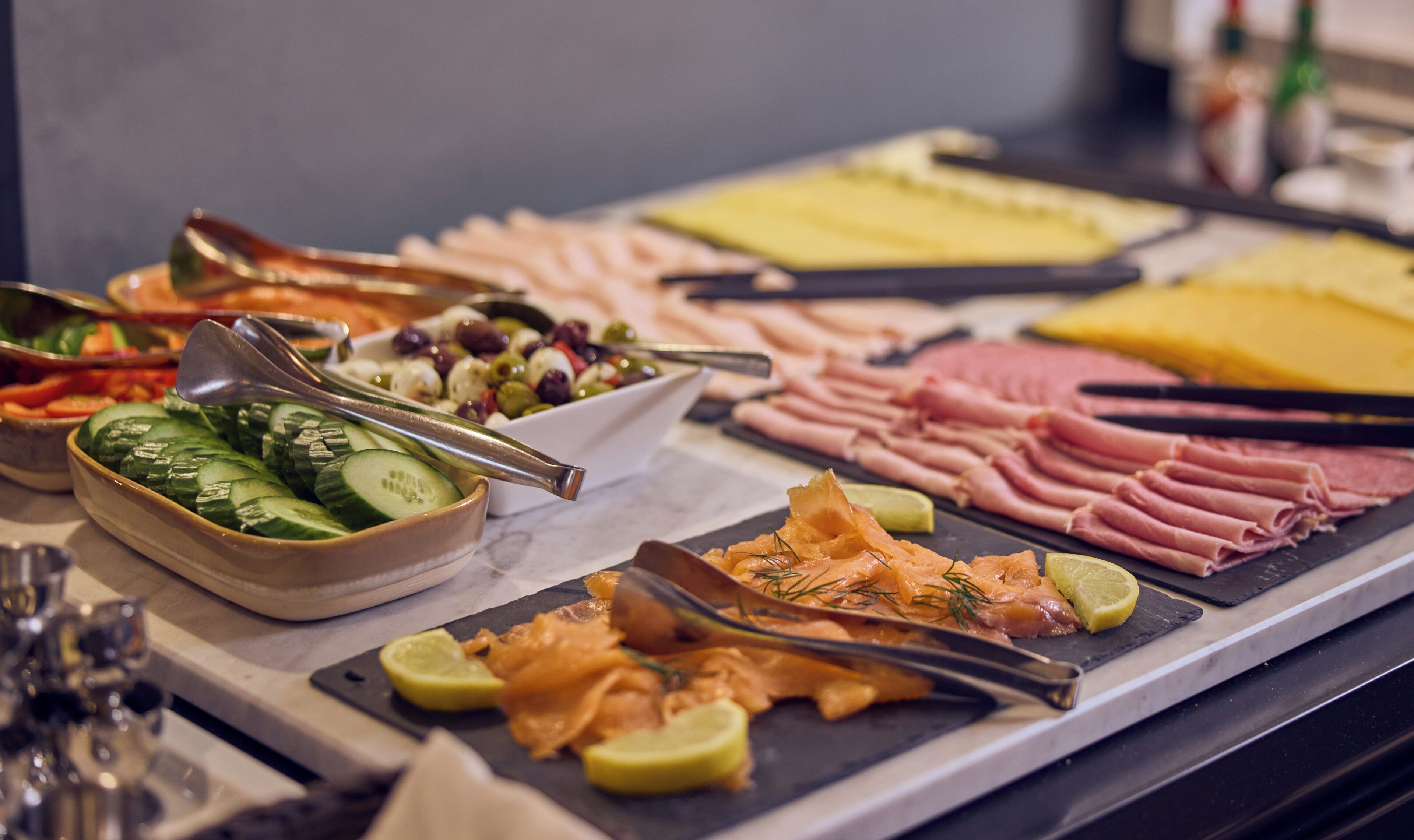 Buffet com diversos alimentos, incluindo pepinos fatiados, azeitonas, presunto e salmão.