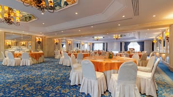 Uma sala de banquetes com mesas redondas e cadeiras brancas, carpete azul, lustres e espelhos grandes.