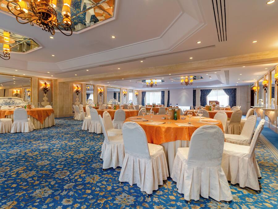 Uma sala de banquetes com mesas redondas e cadeiras brancas, carpete azul, lustres e espelhos grandes.