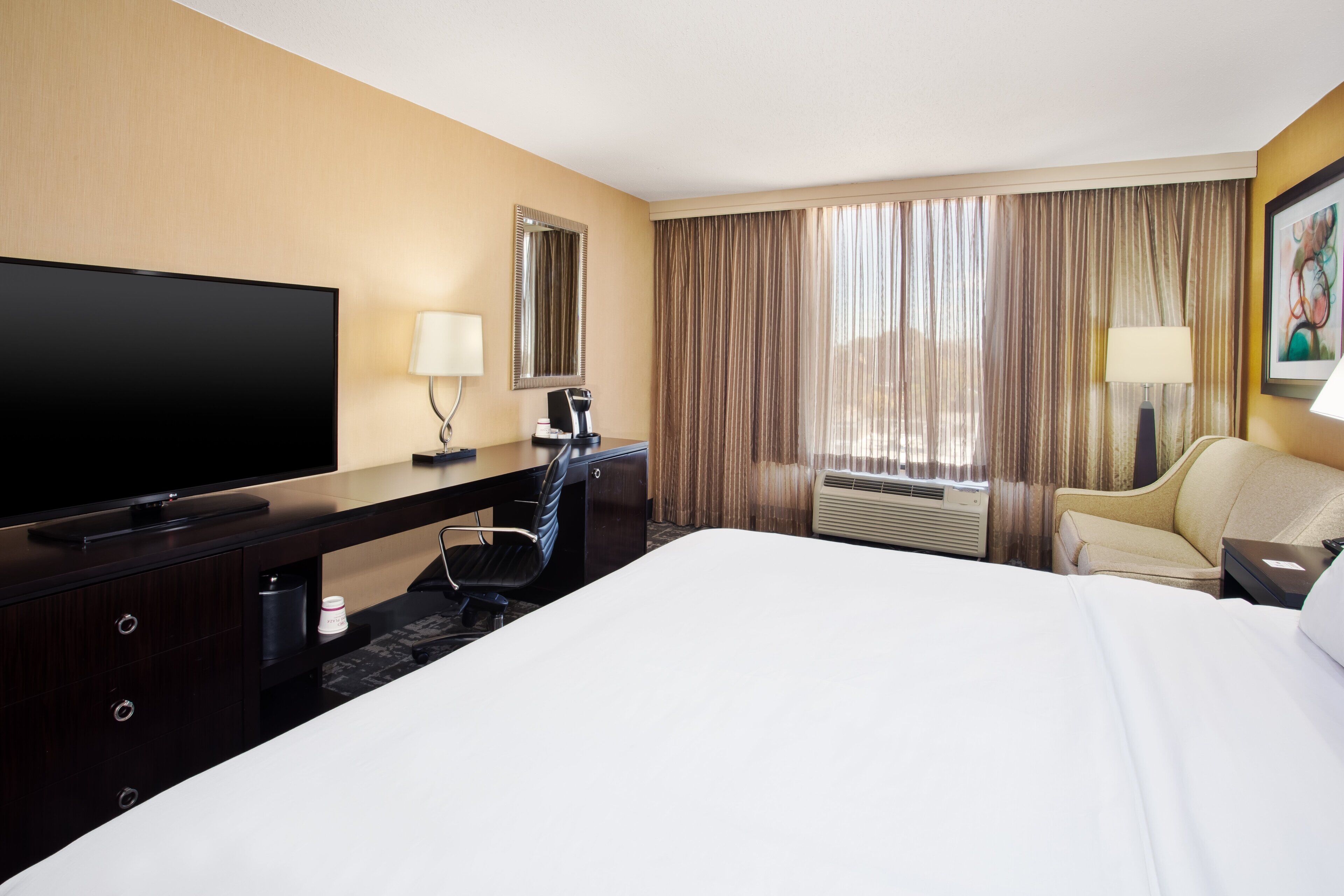 Quarto de hotel com uma cama grande, televisão de tela plana, escrivaninha com cadeira, abajur, cafeteira e janela com cortinas.