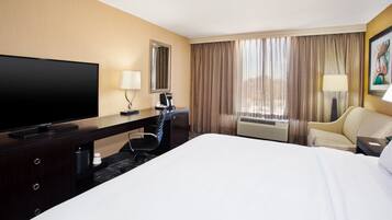 Quarto de hotel com uma cama grande, televisão de tela plana, escrivaninha com cadeira, abajur, cafeteira e janela com cortinas.