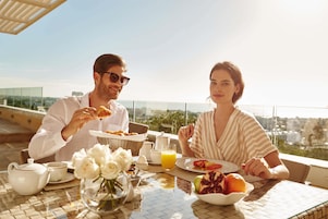 Um casal jantando à mesa com vista para a cidade, com comidas de café da manhã e um vaso de flores.