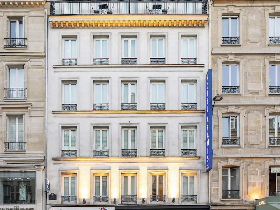 Um hotel de vários andares com estilo arquitetônico europeu clássico, com entrada central com toldo preto, um restaurante visível chamado "Hotel" e uma loja chamada "Réparation Express".