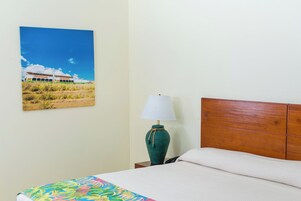 Quarto de hotel com uma cama, mesa de cabeceira com luminária e um quadro na parede retratando um edifício.
